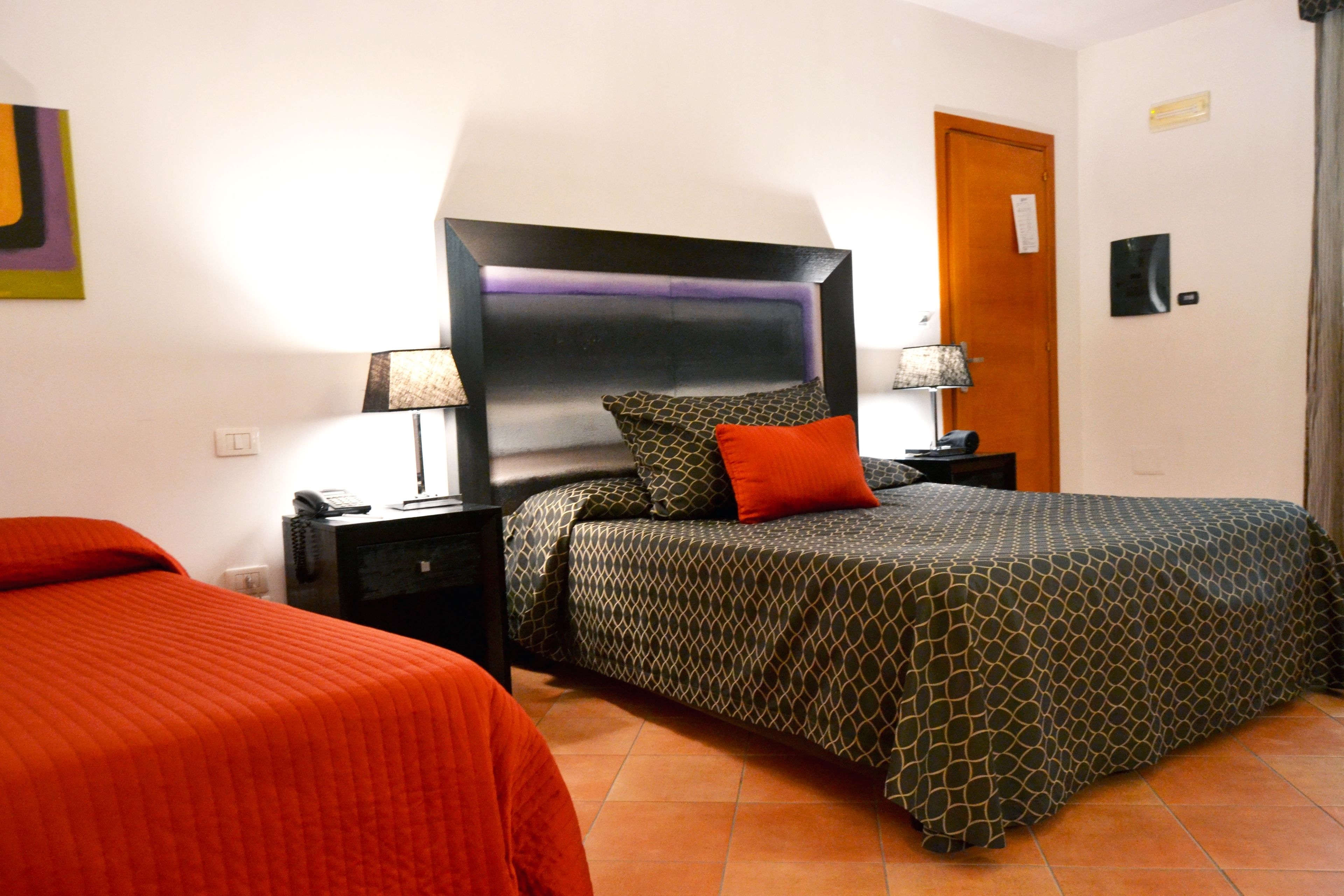 Quarto de hotel com uma cama grande, uma cama menor, uma mesa de cabeceira com abajur e uma porta com um aviso.