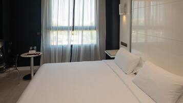 Quarto de hotel com uma cama grande, uma mesinha com uma cadeira, uma janela com cortinas e um espelho na parede.