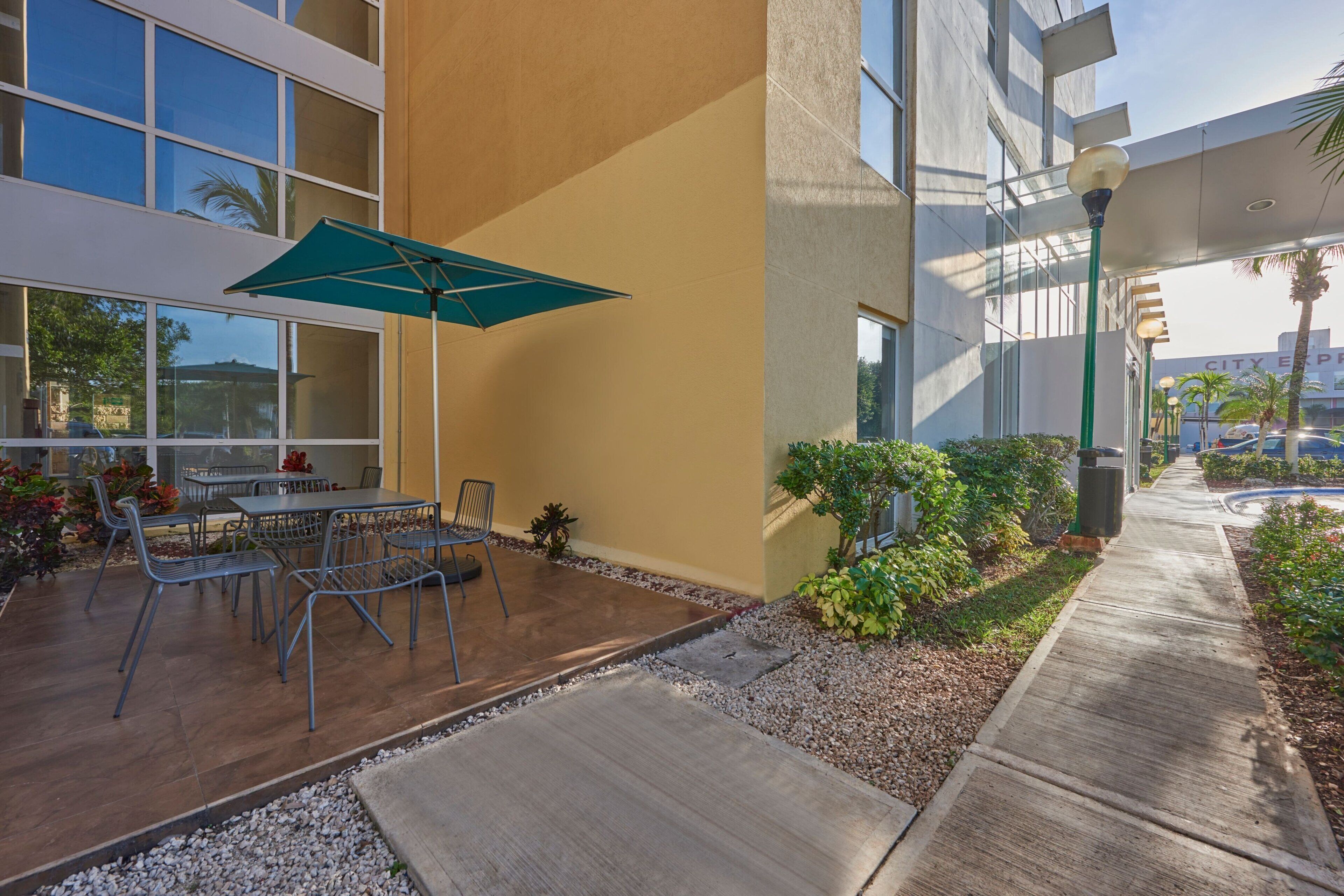 Um pátio externo com mesa e cadeiras, um guarda-sol verde e um caminho que leva a um edifício.