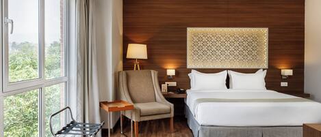 Quarto de hotel com uma janela grande, uma cama, uma cadeira, uma mesa de cabeceira e um painel decorativo na parede.