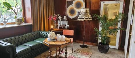 Uma sala de estar com um sofá de couro verde, uma mesa de jantar de madeira com velas, um tapete estampado e uma grande planta em vaso.