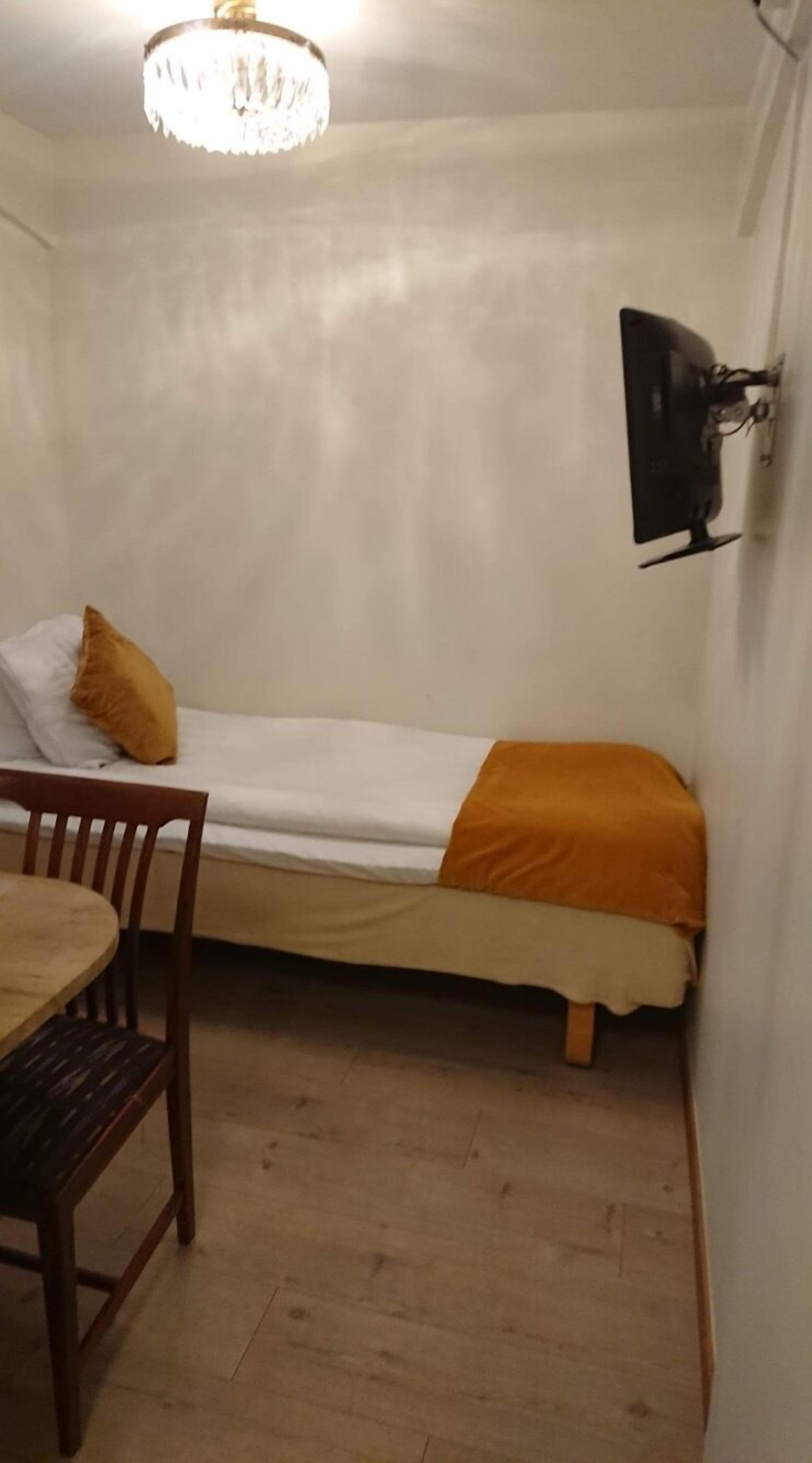 Quarto pequeno com uma cama de solteiro, piso de madeira, uma mesa com cadeiras, TV fixada na parede e um candelabro.