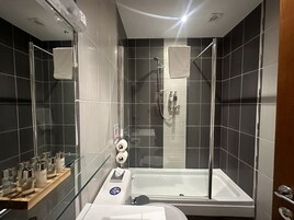 Casa de banho moderna com chuveiro em cabine de vidro, banheira branca e prateleira de vidro com artigos de higiene pessoal.