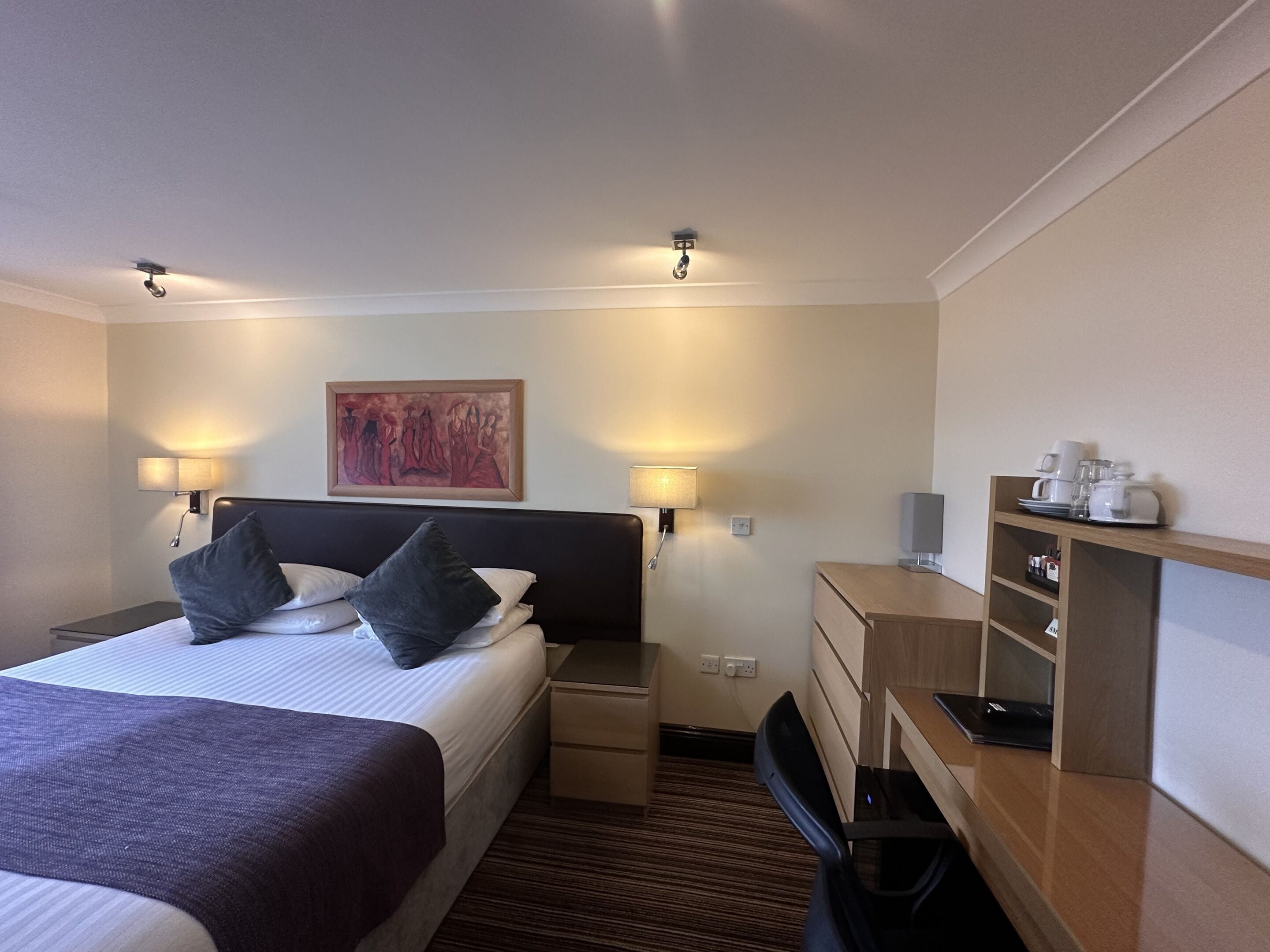 Quarto de hotel com cama, abajures, escrivaninha com computador, cadeira e um quadro na parede.