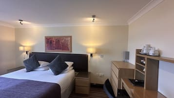 Quarto de hotel com cama, abajures, escrivaninha com computador, cadeira e um quadro na parede.