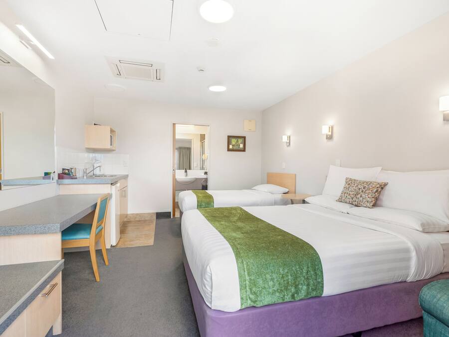 Quarto de hotel com escrivaninha, duas camas, banheiro e corredor.