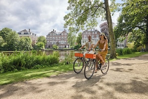Duas pessoas de bicicleta com cestas, um lago e edifícios históricos ao fundo.