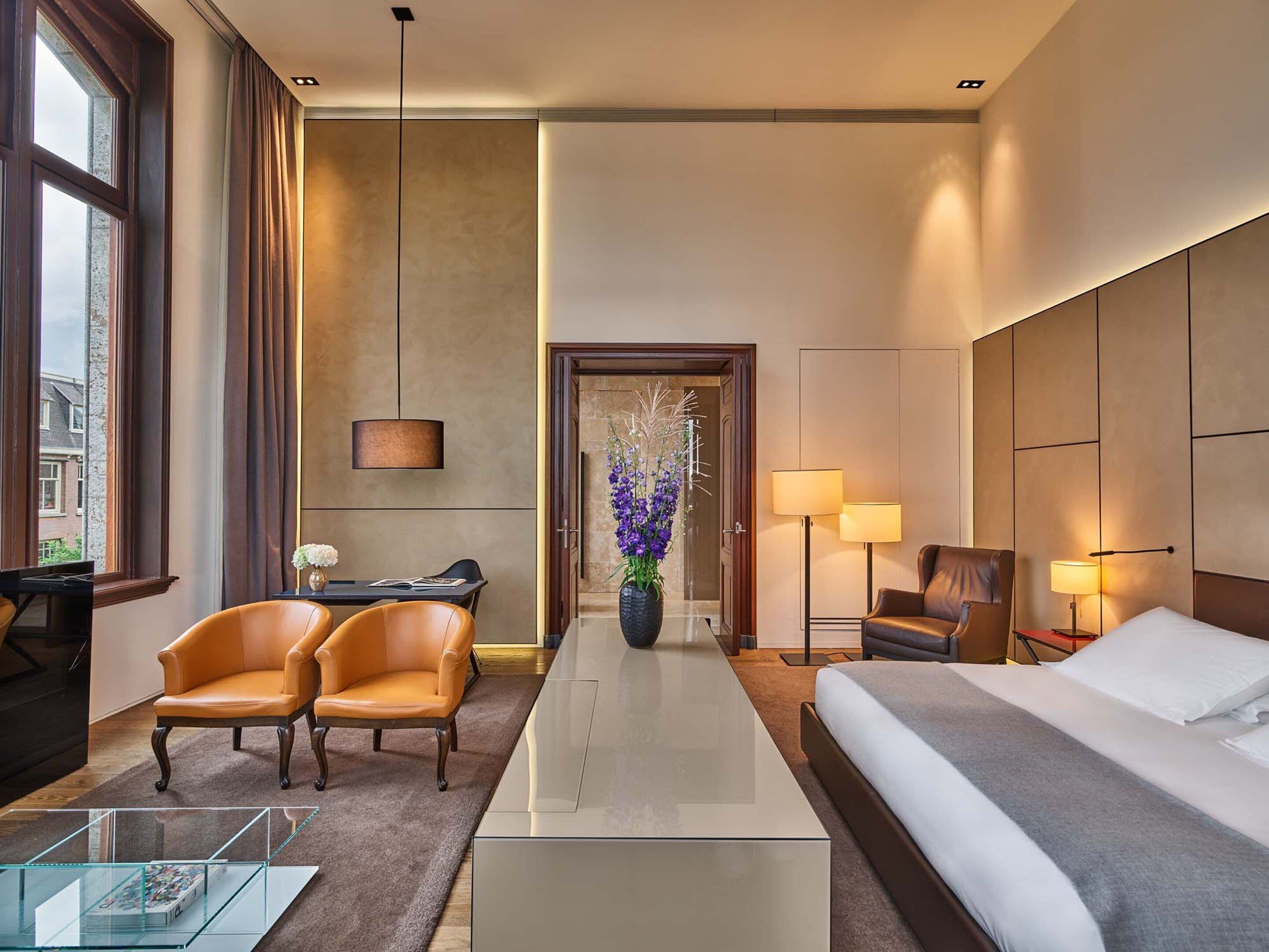 Quarto de hotel com uma cama, duas cadeiras, uma escrivaninha, um vaso com flores e um abajur.