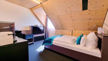 Quarto de hotel moderno com teto de madeira, cama com roupa de cama branca e azul, uma escrivaninha com computador e uma televisão.