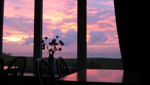 Mesa com cadeiras e um vaso de flores, com o céu do pôr do sol ao fundo, visto através de uma janela.