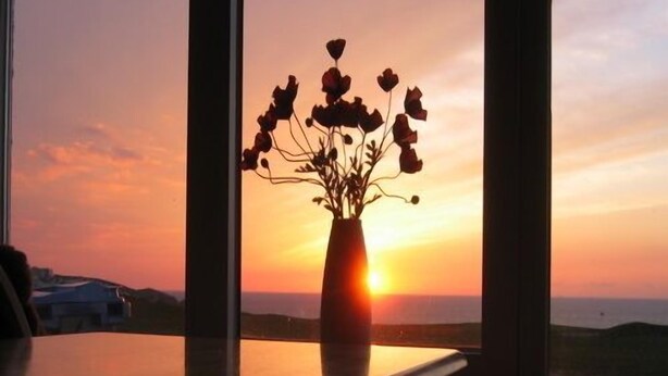 Vista do pôr do sol através de uma janela, com um vaso de flores sobre uma mesa.