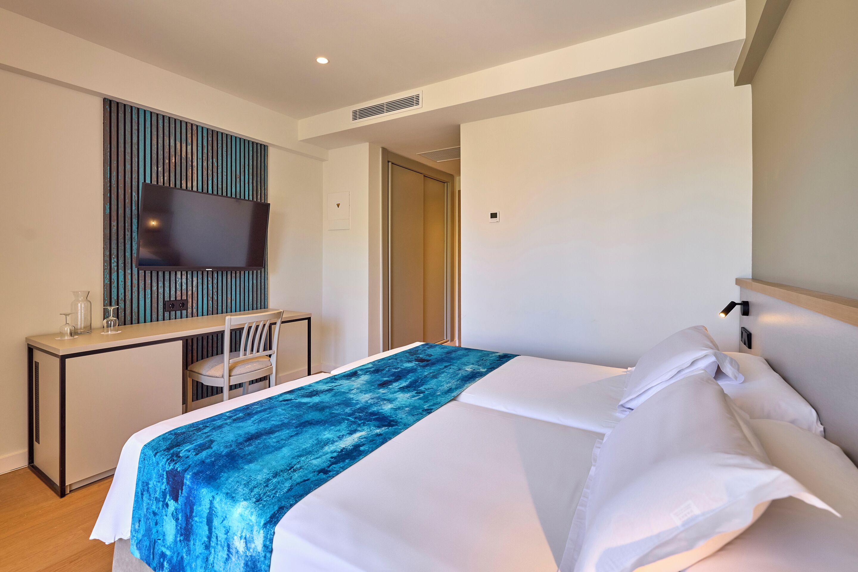 Quarto de hotel com cama, escrivaninha, cadeira, TV fixada na parede e porta de acesso a outro cômodo.