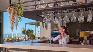 Uma área de bar com vista para a praia, banquetas, copos de vinho pendurados e um bartender.