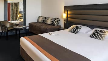 Quarto de hotel com uma cama grande, um sofá, uma poltrona, uma mesinha e um espelho.