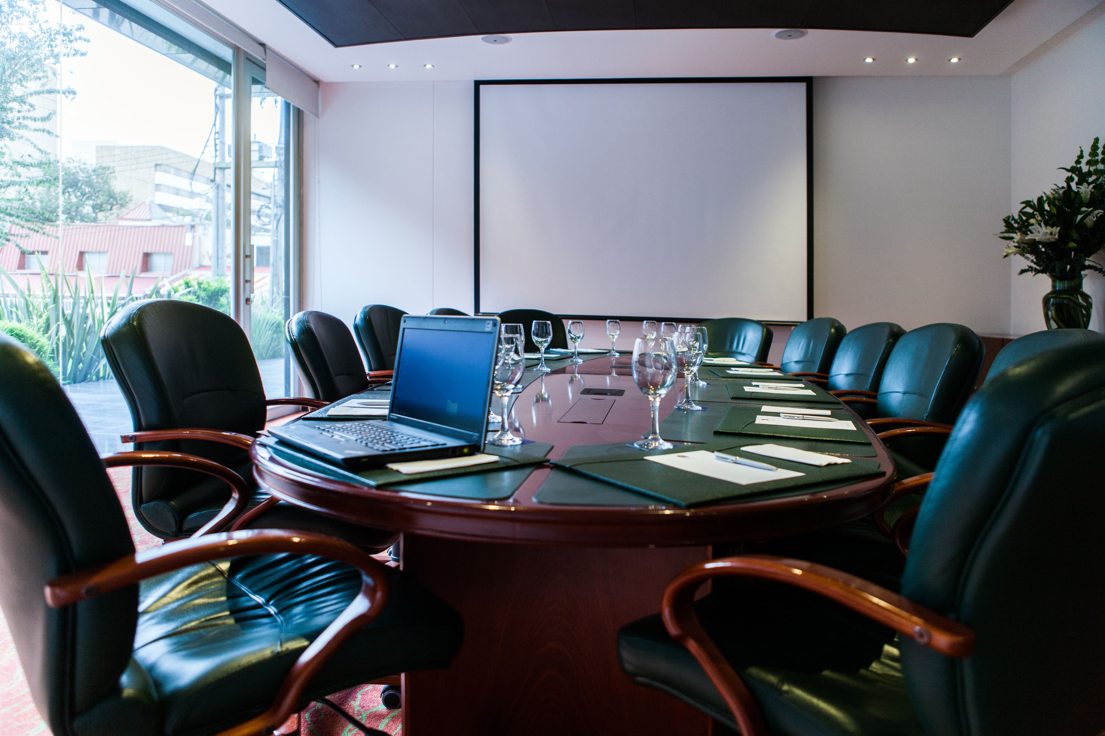 Sala de conferências com mesa redonda, cadeiras de couro preto, um laptop e copos de vinho.