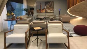 Uma sala de estar moderna com um conjunto de sofás, uma mesa de centro e um puff vermelho. Há quadros abstratos na parede e uma pequena mesa com objetos decorativos.