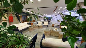 Uma área de estar interna moderna, com sofás, cadeiras e mesas redondas, cercada por plantas em vasos.
