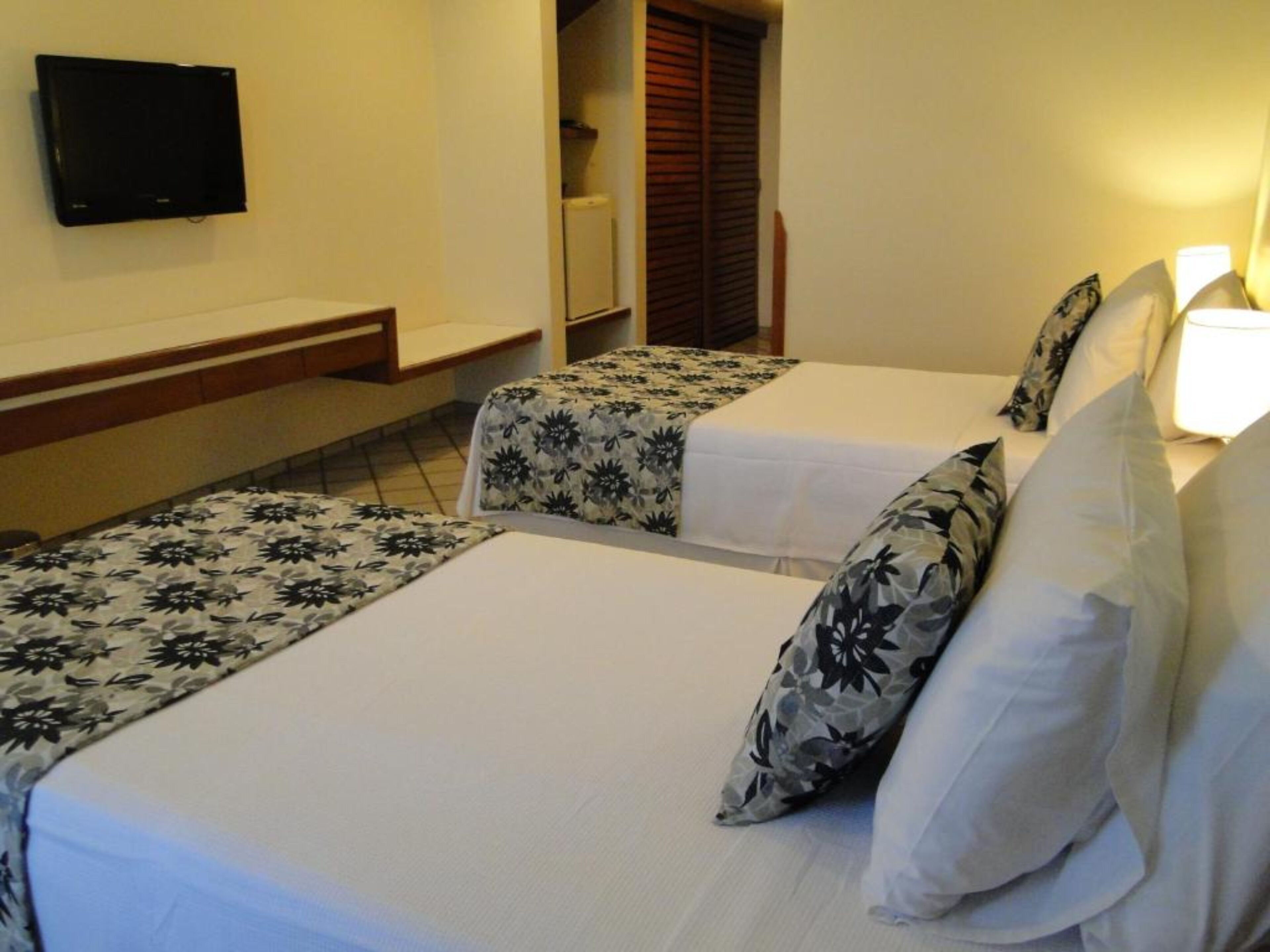 Quarto de hotel com duas camas, cada uma com colchas florais e lençóis brancos. Há uma televisão de tela plana na parede e uma mesa de cabeceira com abajur.