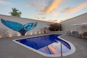 Piscina com um mural de uma cauda de baleia, rodeada por cadeiras de espreguiçadeira e um guarda-sol.