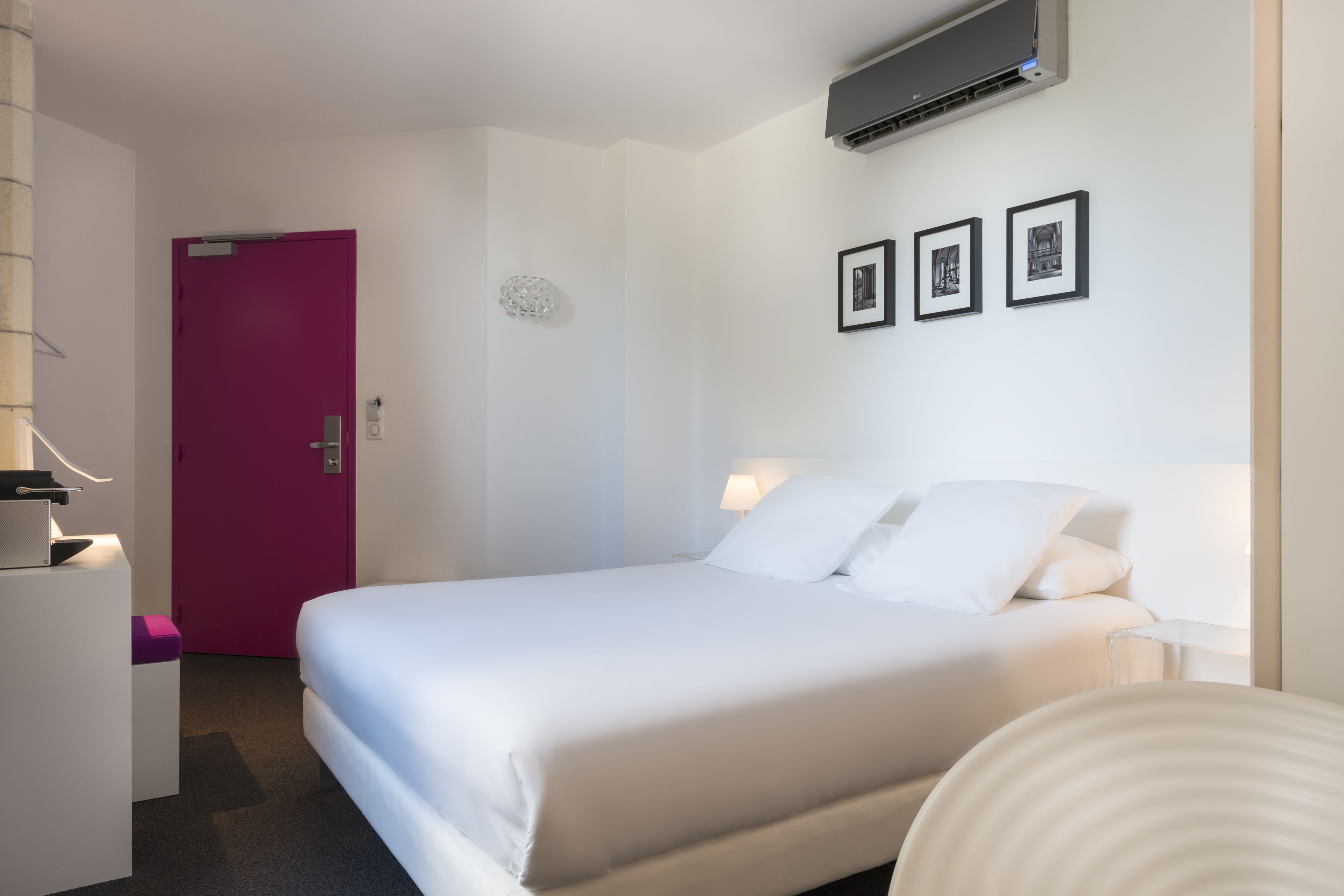 Quarto de hotel com cama, escrivaninha, cadeira, porta e três quadros na parede.