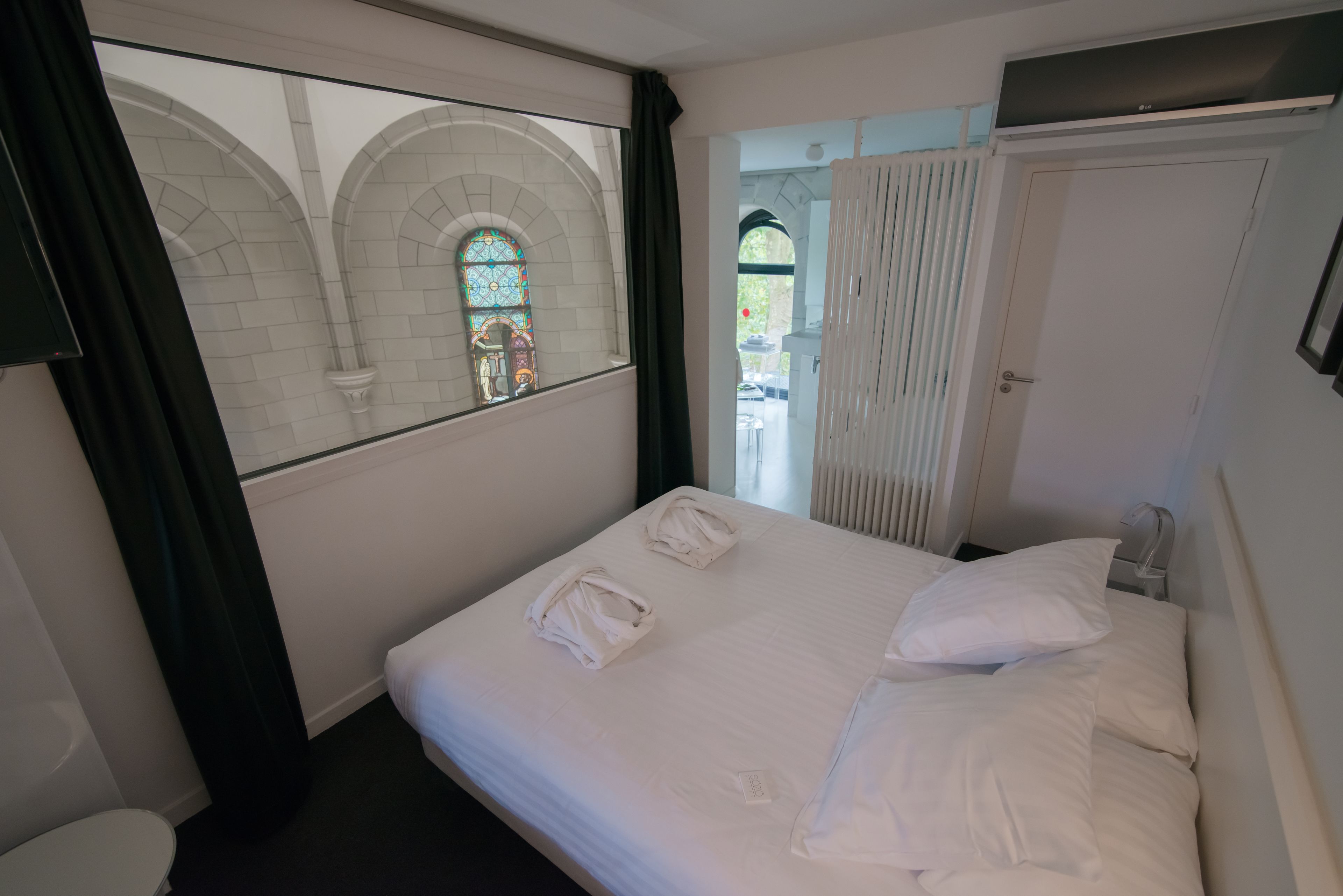 Quarto de hotel com uma cama, uma janela com vista para o interior de um edifício, uma porta branca e um radiador branco.