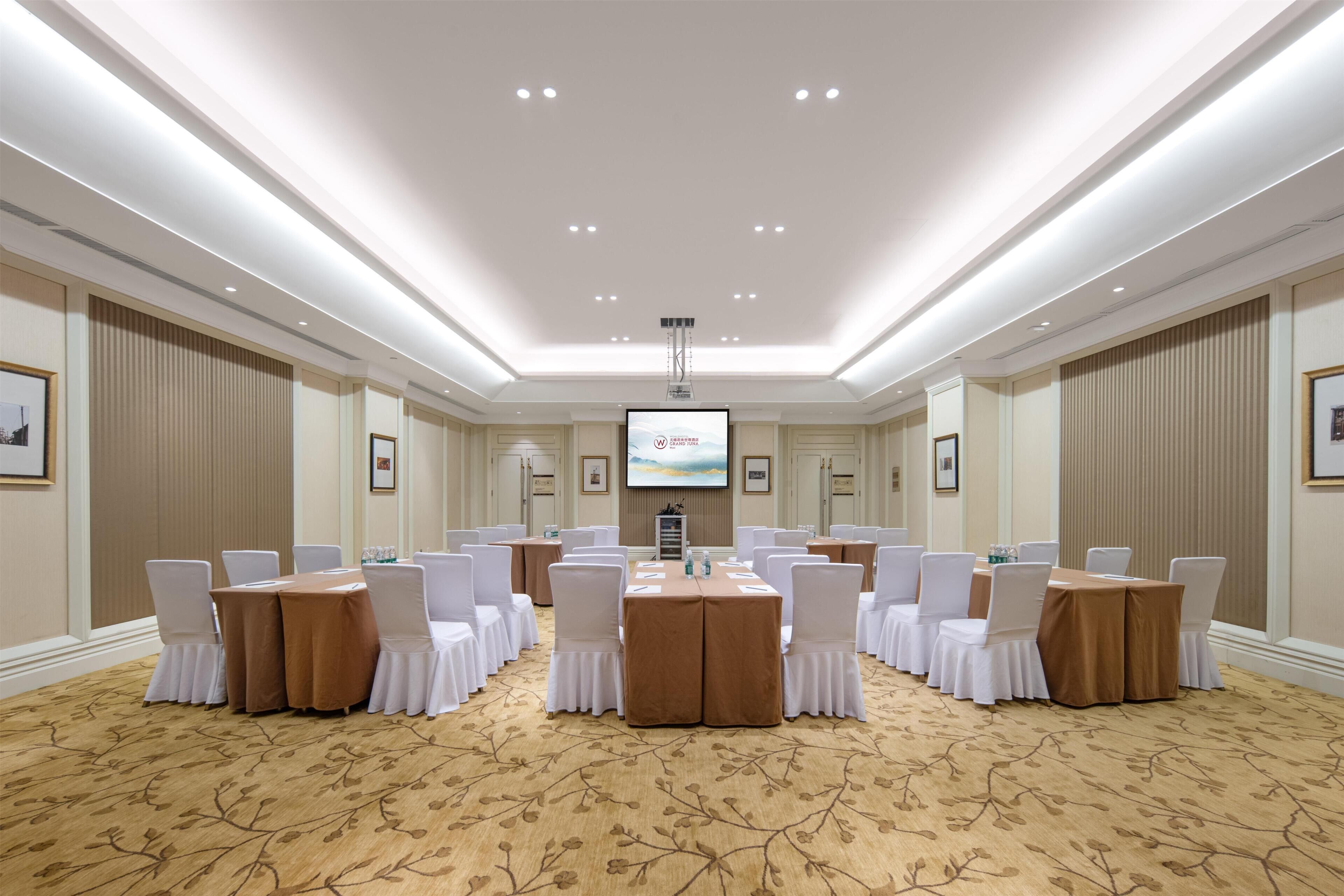 Sala de conferências com tela grande, diversas mesas e cadeiras.