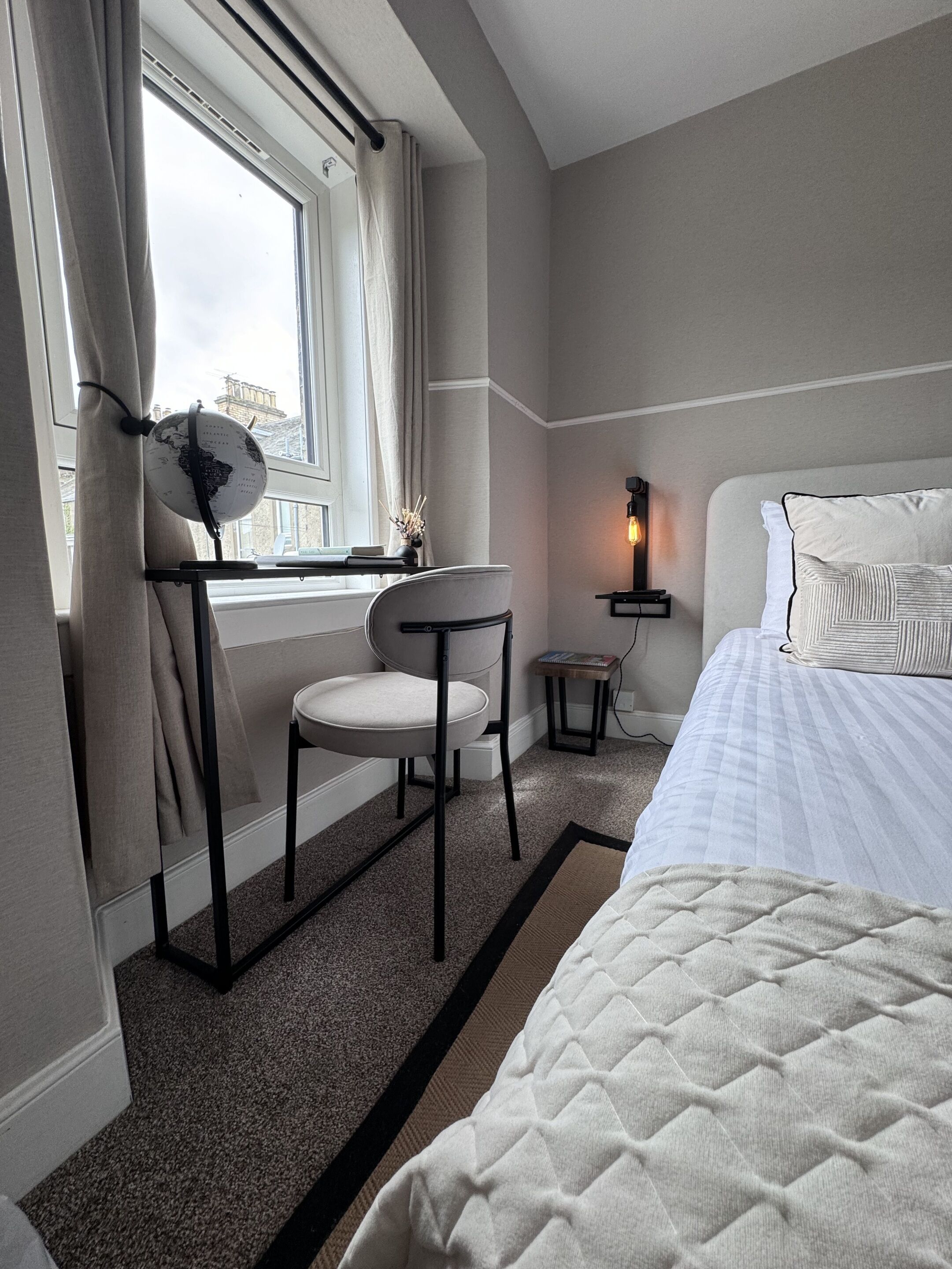 Quarto de hotel com uma cama, uma escrivaninha, uma cadeira, uma mesinha, um globo e uma mesa de cabeceira com uma luminária.