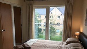 Quarto com uma janela ampla, cama com roupa de cama bege, mesa de cabeceira com abajur e vista para prédios.