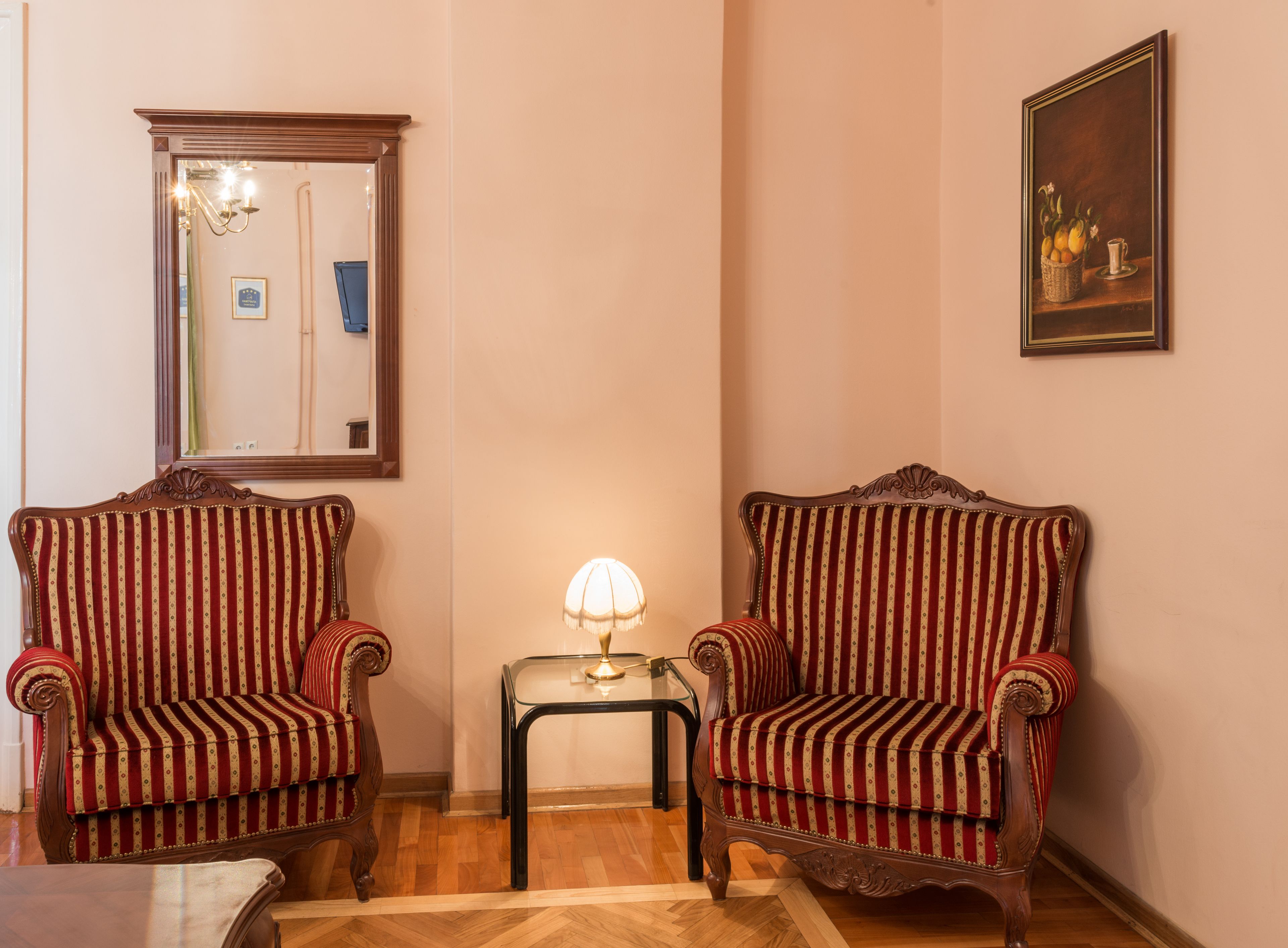 Um quarto com duas poltronas elaboradas, uma mesinha de centro com abajur, espelho e um quadro emoldurado na parede.
