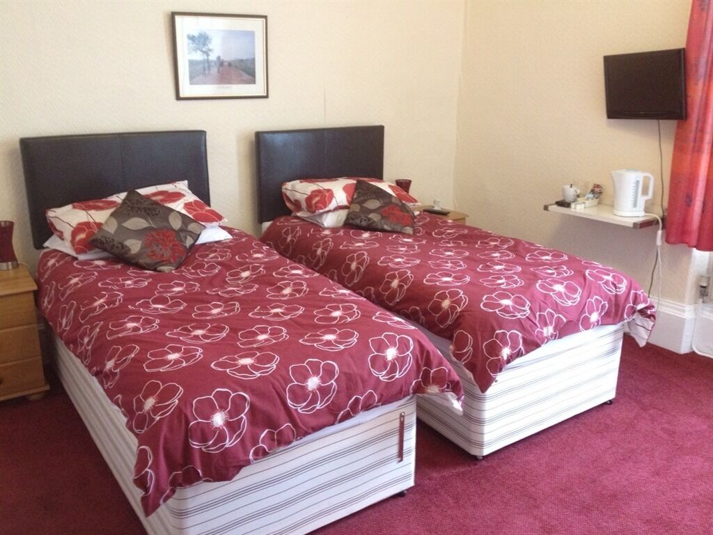 Quarto de hotel com duas camas, cada uma com colcha e almofadas estampadas com motivos florais. Há uma pequena mesa de cabeceira com uma luminária, um quadro pendurado na parede e uma prateleira com uma chaleira e outros objetos.