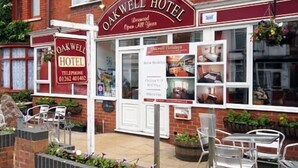 Exterior do Hotel Oakwell com fachada tradicional, número de telefone visível e uma pequena área externa com cadeiras e mesas de metal.