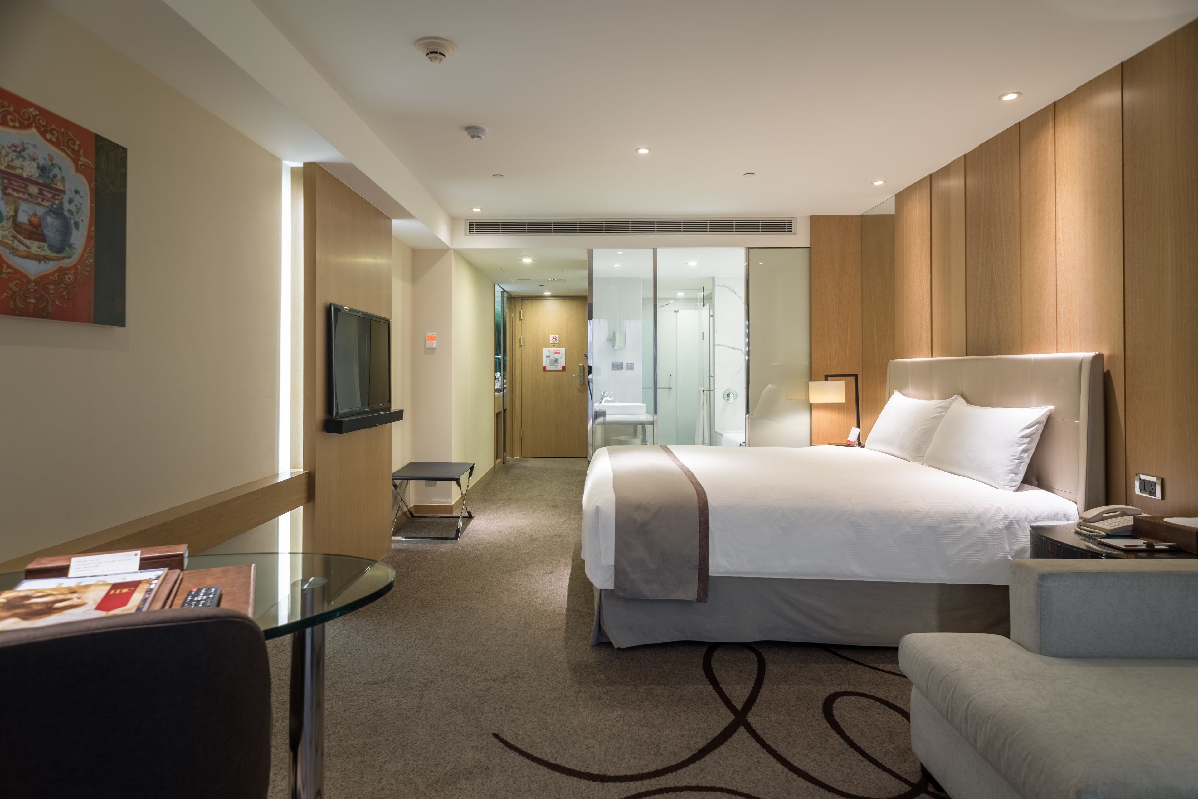 Quarto de hotel com cama, escrivaninha, cadeira, mesinha de vidro e um quadro na parede.