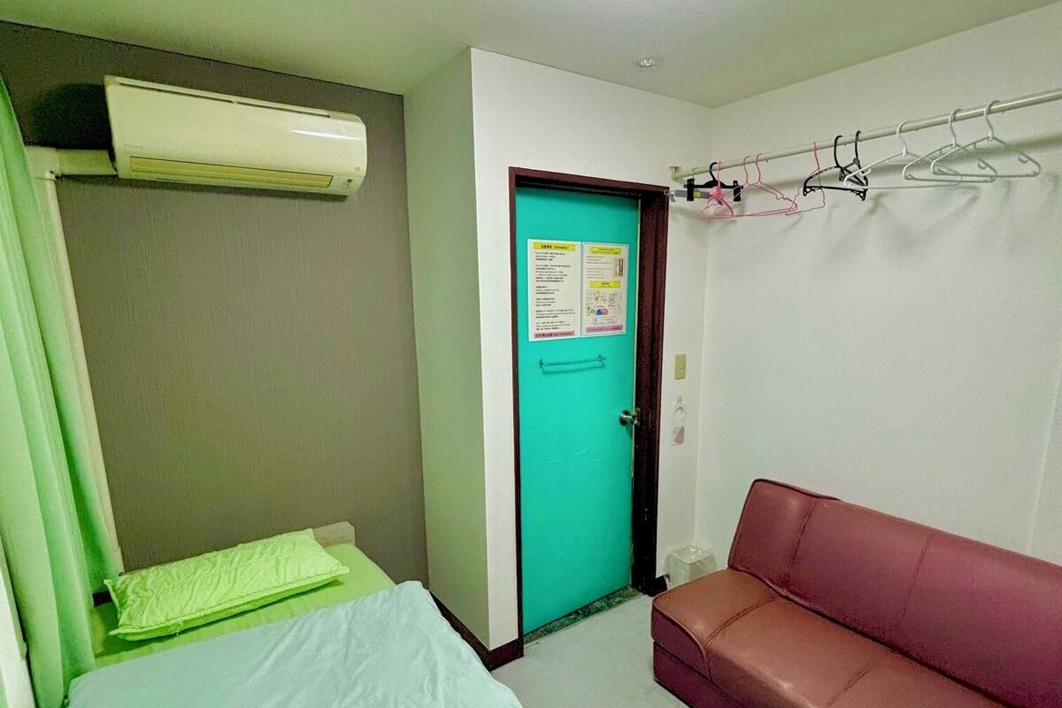 Um quarto com cama, sofá, aparelho de ar condicionado, porta com aviso e um porta-roupas.