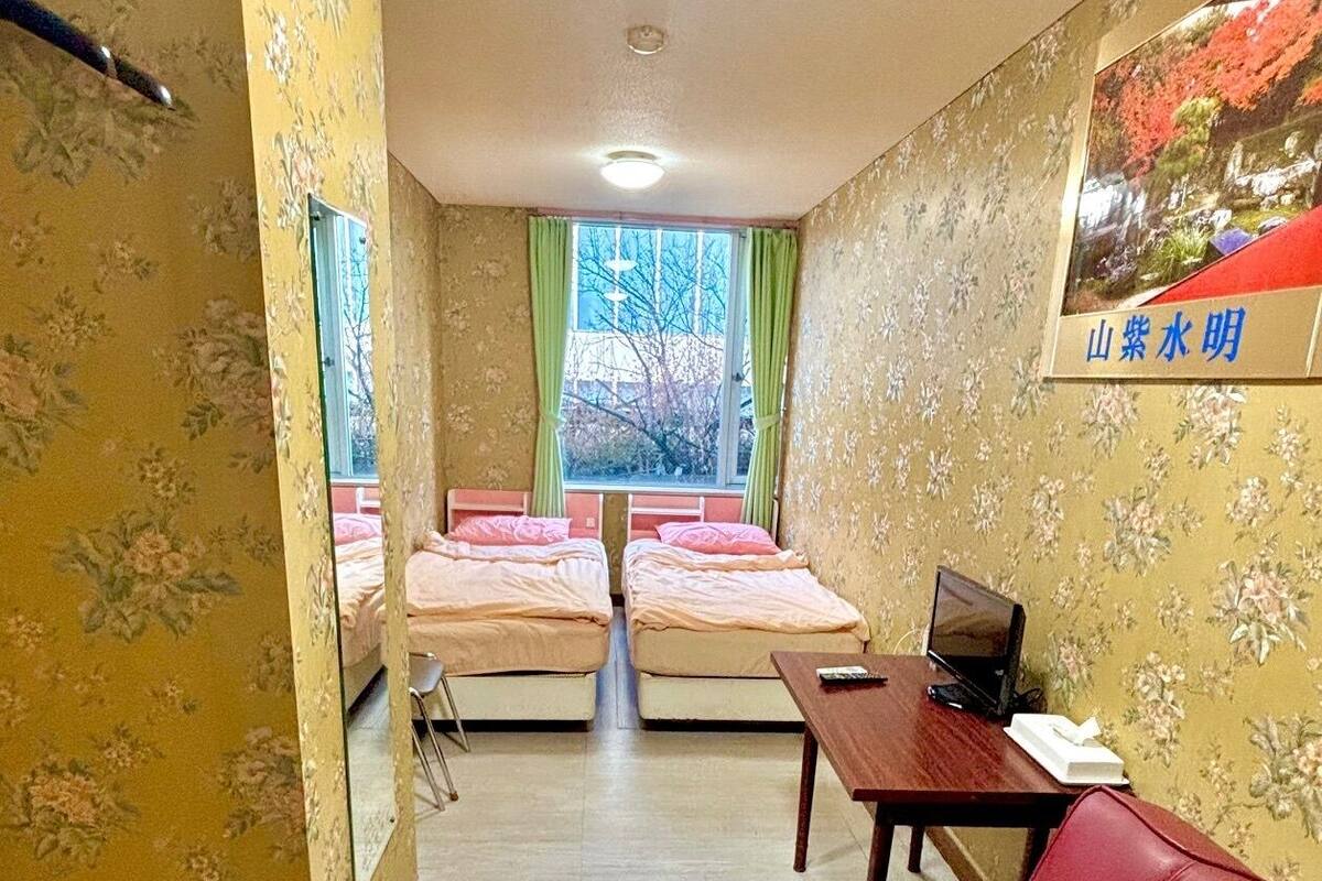 Quarto de hotel com duas camas, um sofá vermelho, uma escrivaninha com um laptop e uma janela com cortinas verdes.
