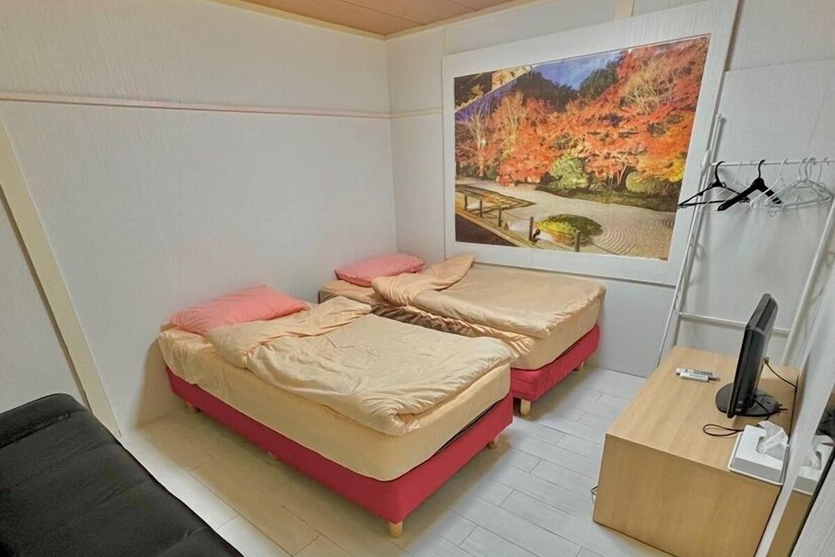 Quarto pequeno e compacto com duas camas, uma escrivaninha com computador e janela com vista para árvores de outono.