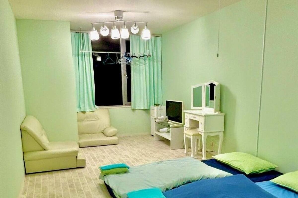 Quarto com duas camas, uma cadeira, escrivaninha com computador e janela com cortinas.