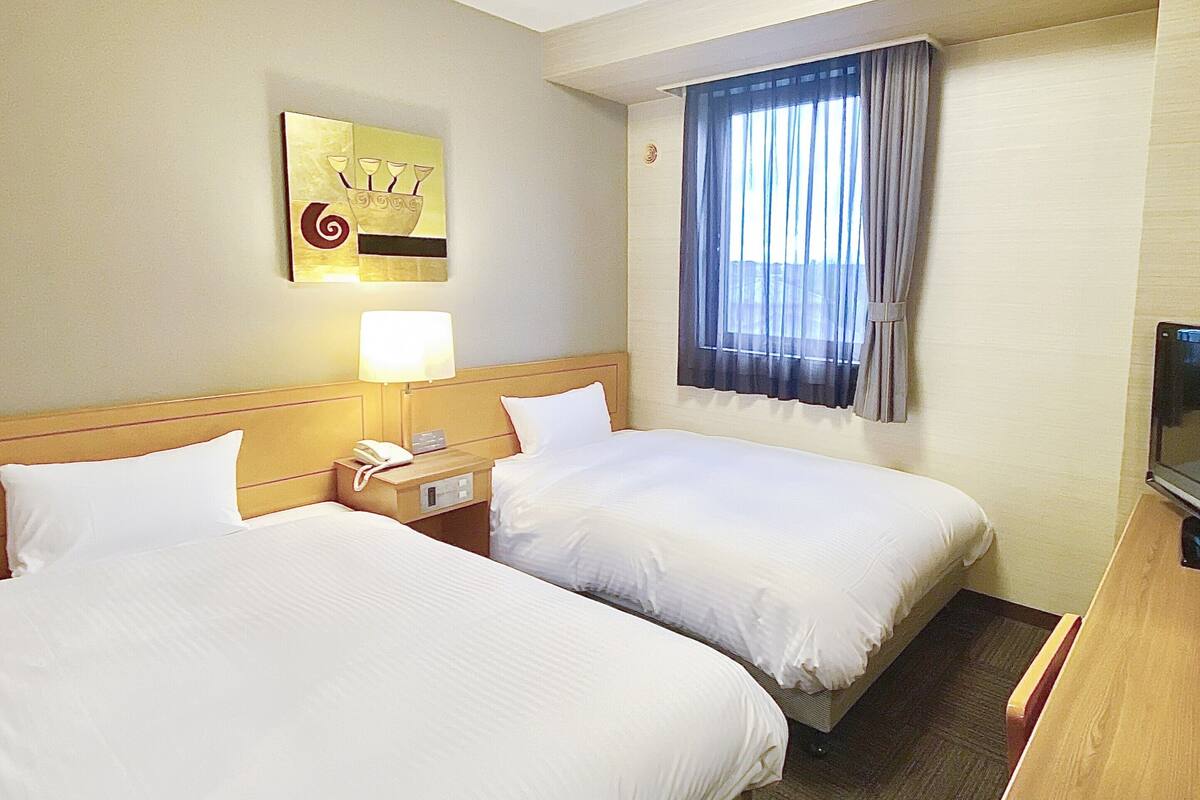 Quarto de hotel com duas camas, mesa de cabeceira com abajur, televisão e janela com cortinas.