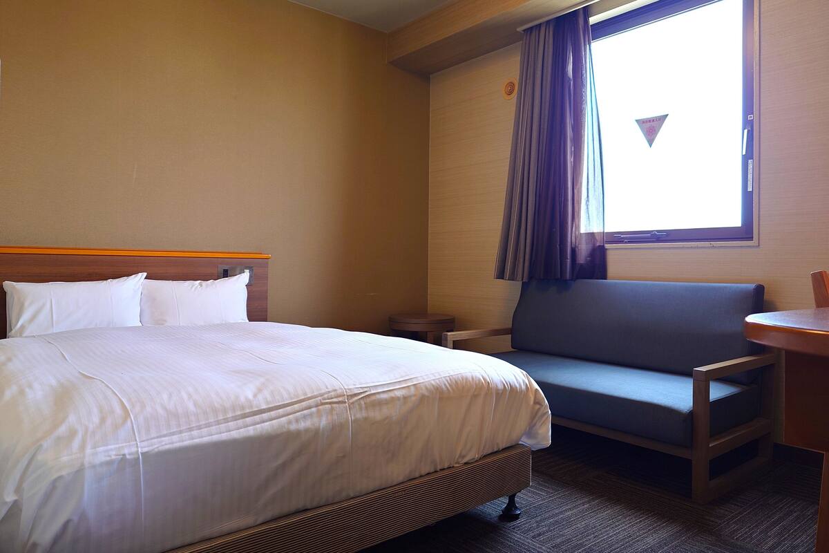 Quarto de hotel com uma cama, uma mesinha, uma cadeira e uma janela com cortinas.