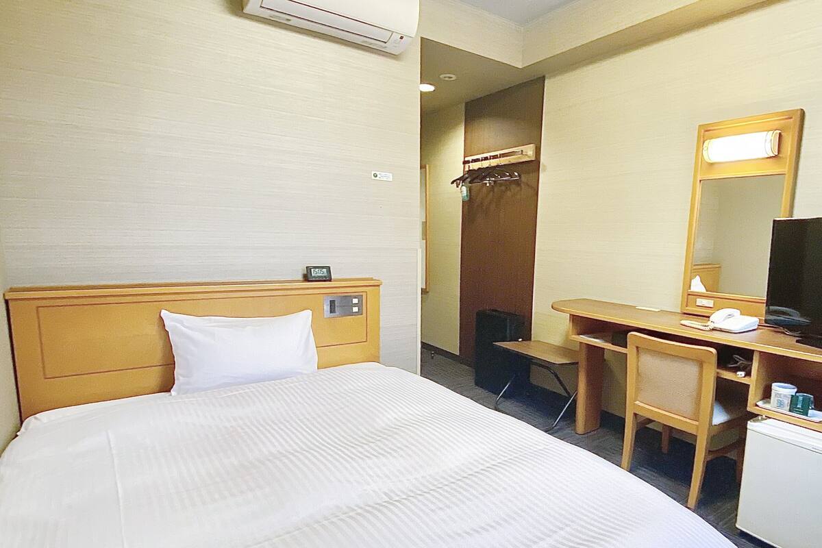 Quarto de hotel com cama, escrivaninha, cadeira, espelho e um pequeno frigorífico.