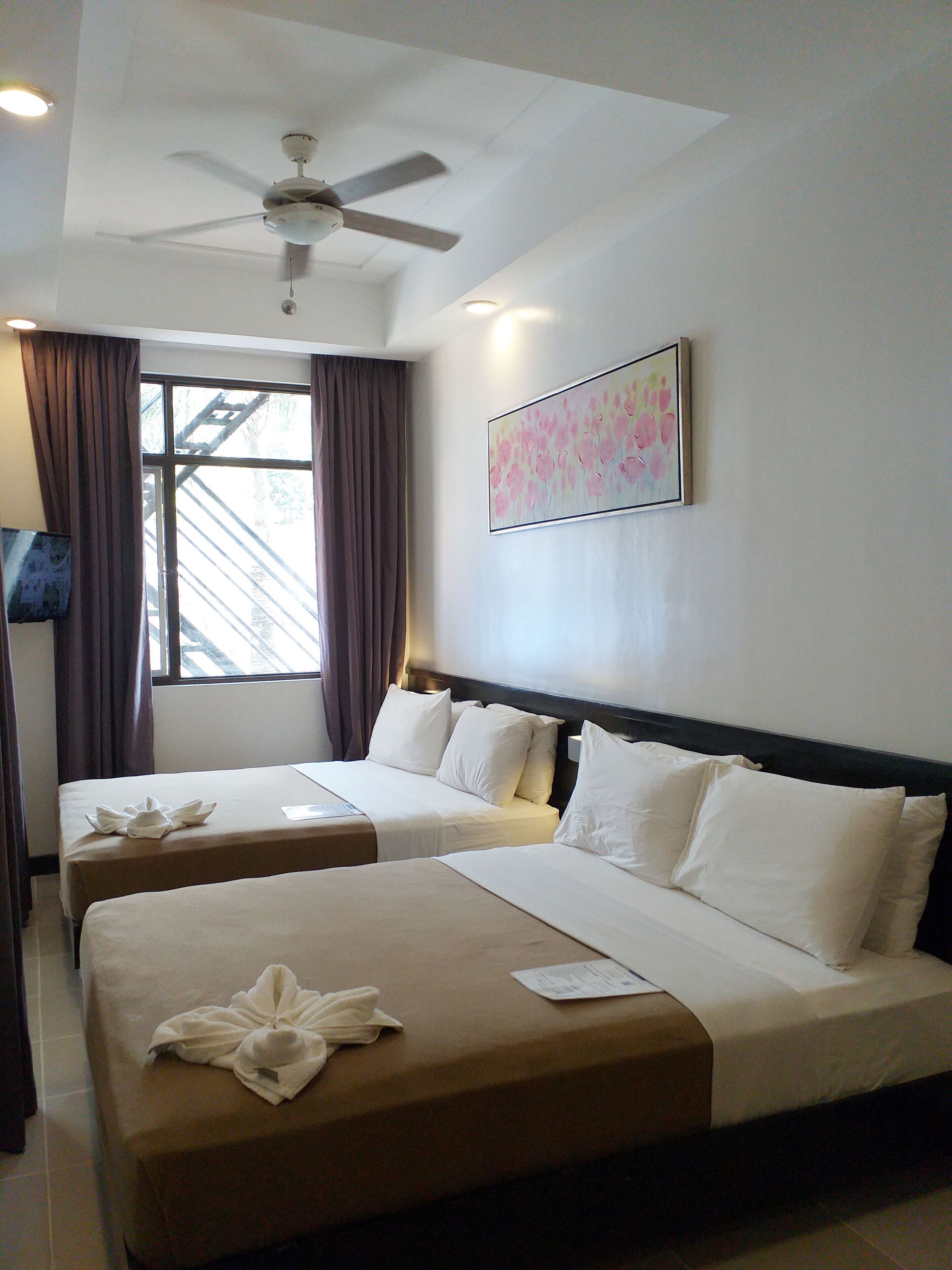 Quarto de hotel com duas camas, um ventilador de teto, janela com cortinas e um quadro na parede.