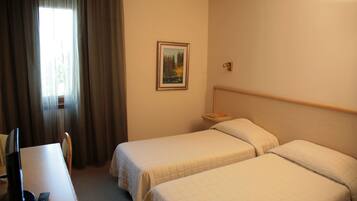 Quarto de hotel com duas camas, uma escrivaninha com controle remoto, uma cadeira, um quadro na parede e uma janela com cortinas.