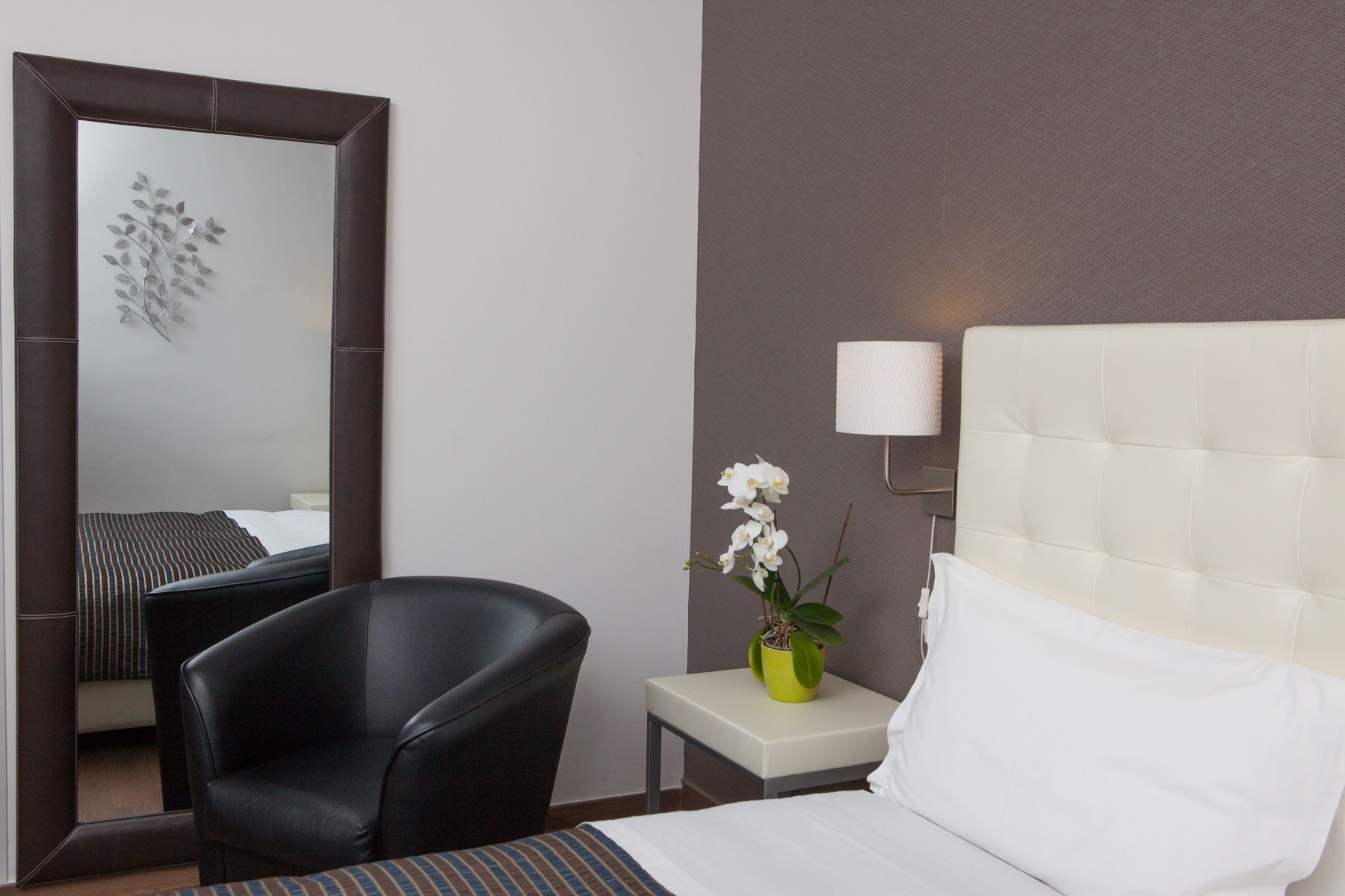 Quarto de hotel com cama, uma cadeira, mesa de cabeceira com abajur e espelho com um ornamento na parede.