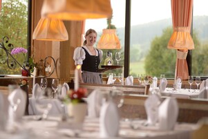 Restaurante com mesas preparadas para refeições, um garçom atendendo aos clientes e uma vista panorâmica pela janela.