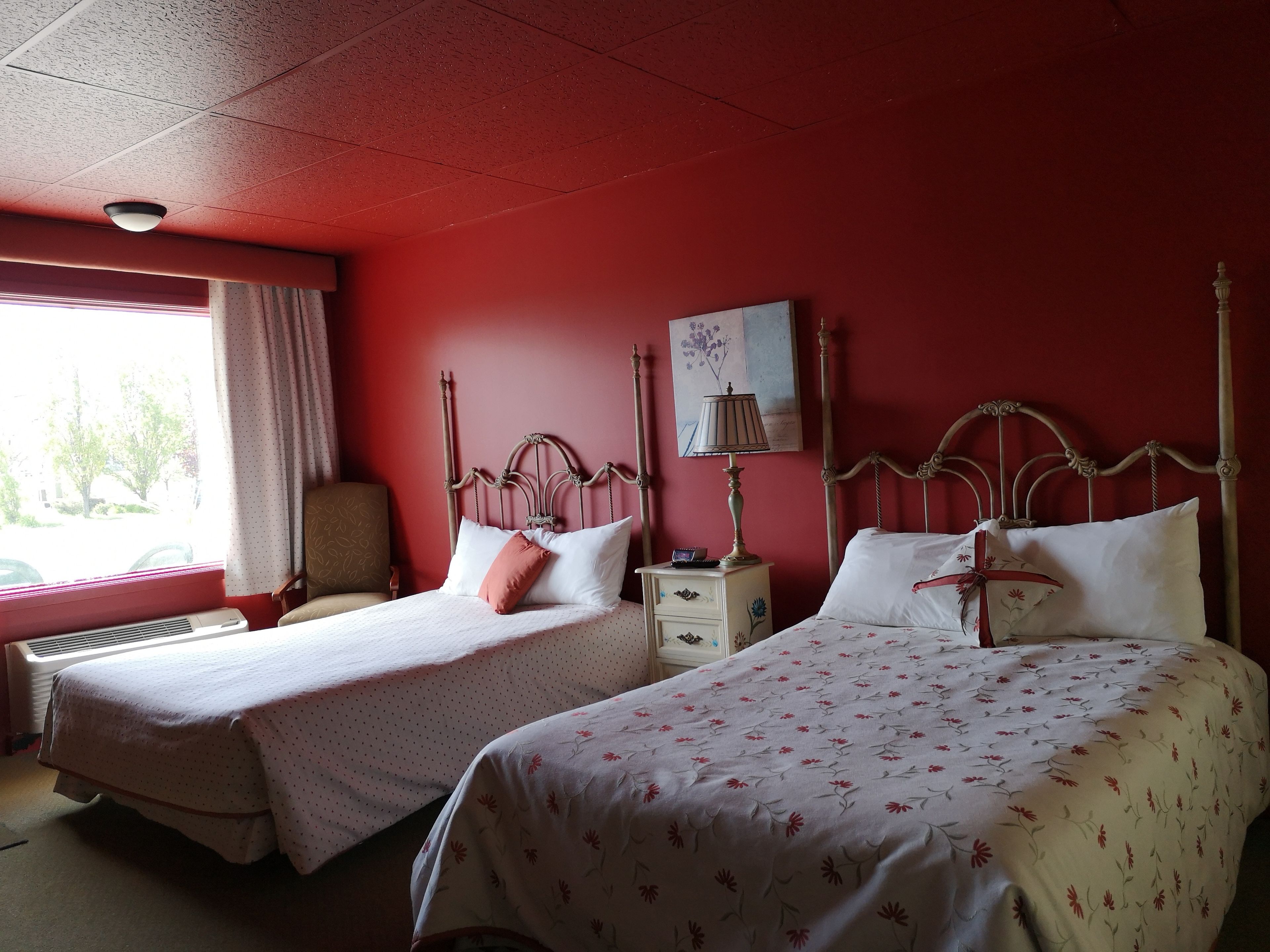 Quarto de hotel com duas camas, janela com cortinas, uma cadeira, mesa de cabeceira com abajur e uma obra de arte na parede.