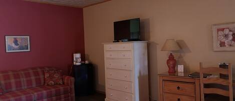 Quarto de hotel com cama, sofá vermelho, penteadeira, televisão sobre a penteadeira, abajur e uma pequena mesa com cadeira.