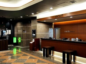 Uma área de recepção de hotel com balcão de madeira, mesa de atendimento e placas de sinalização.