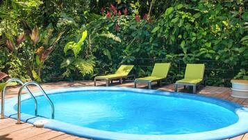 Piscina externa com deck de madeira, cercada por vegetação exuberante e três espreguiçadeiras verdes.