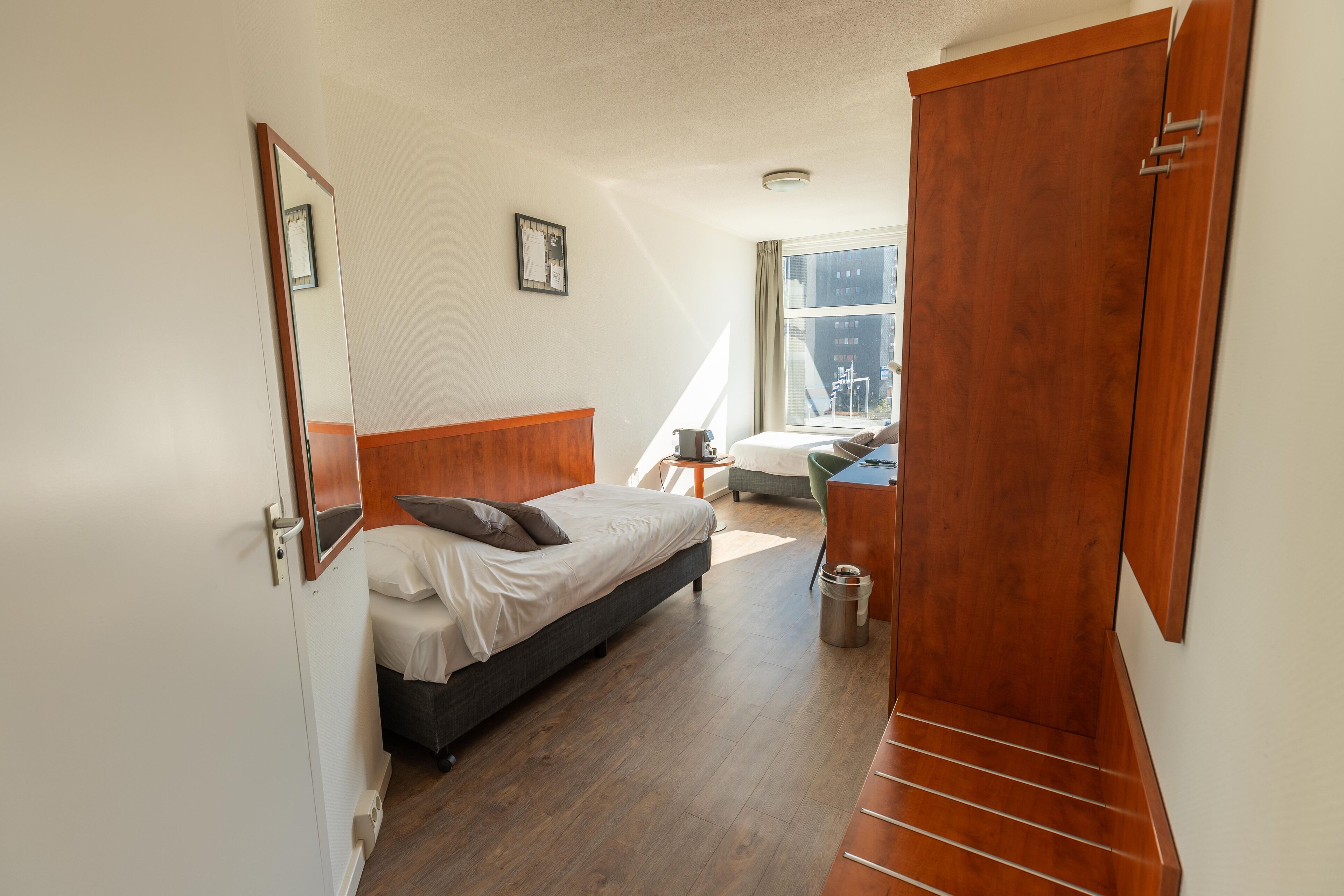 Quarto com cama, guarda-roupa de madeira, escrivaninha e janela com vista para edifícios.