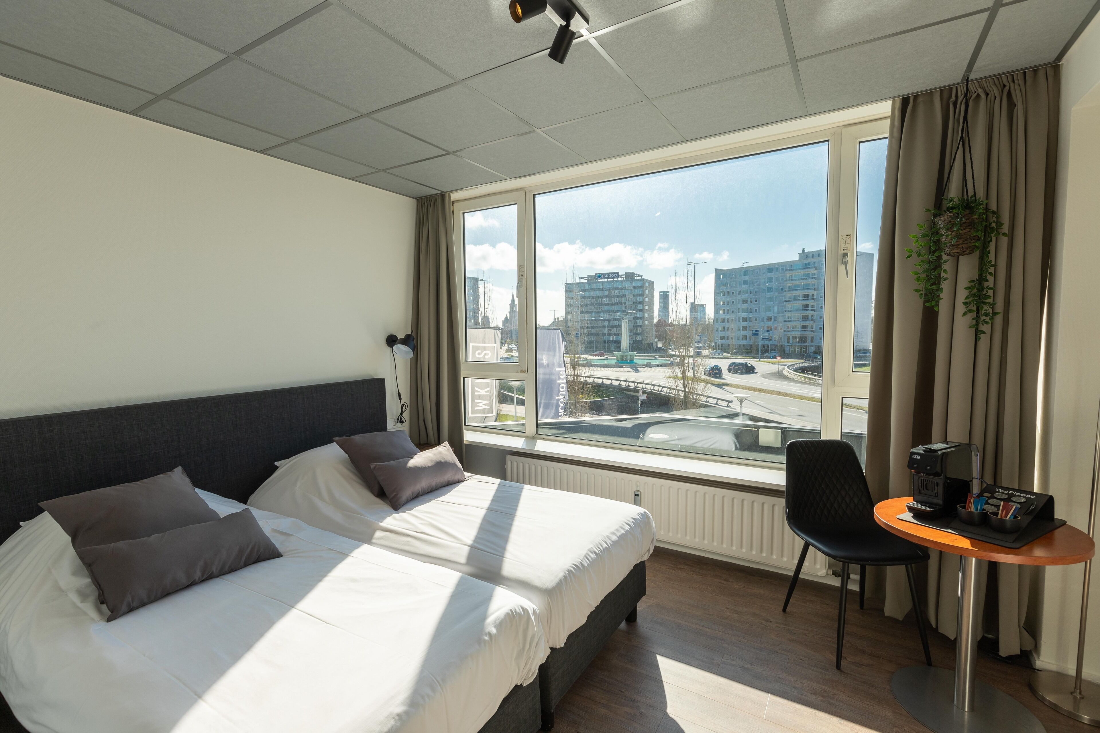 Quarto de hotel moderno com uma cama grande, uma mesinha com cafeteira, uma cadeira e vista da cidade por uma janela ampla.