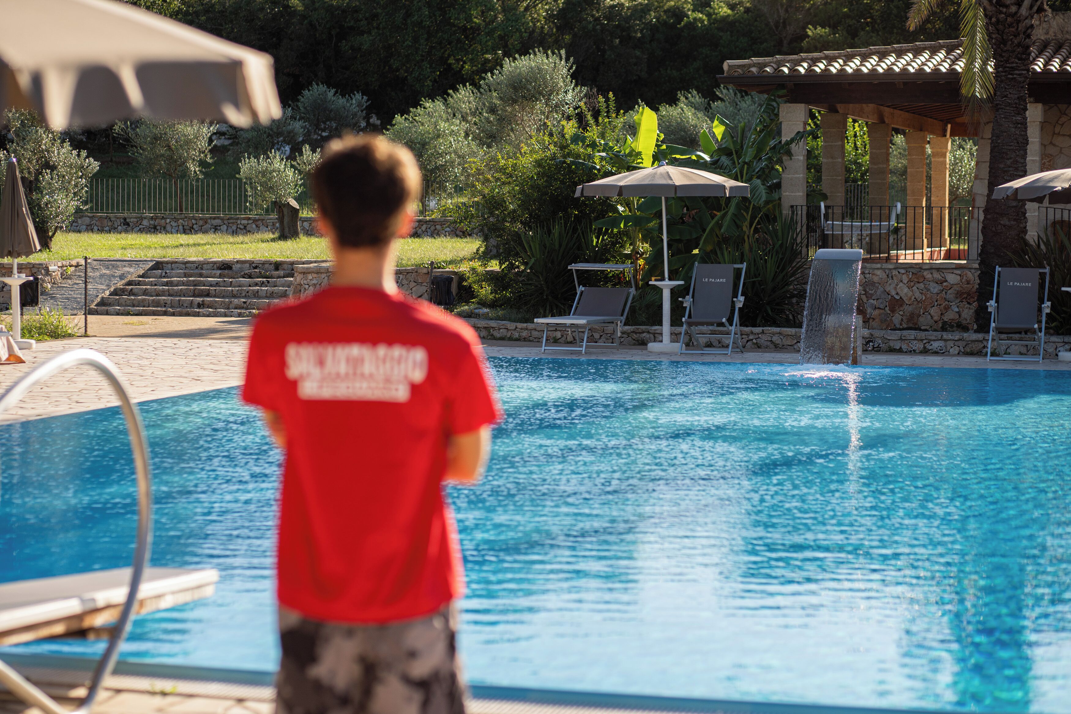 Uma área de piscina com espreguiçadeiras e guarda-sóis, cercada por vegetação e um muro de pedra.